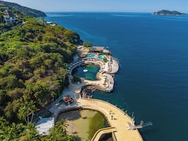 Vista aérea de um resort à beira-mar, com piscina central, área de praia e montanhas ao fundo.