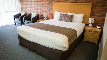 Quarto de hotel com uma cama grande, cabeceira de madeira, parede de tijolos, escrivaninha com abajur e uma cadeira.