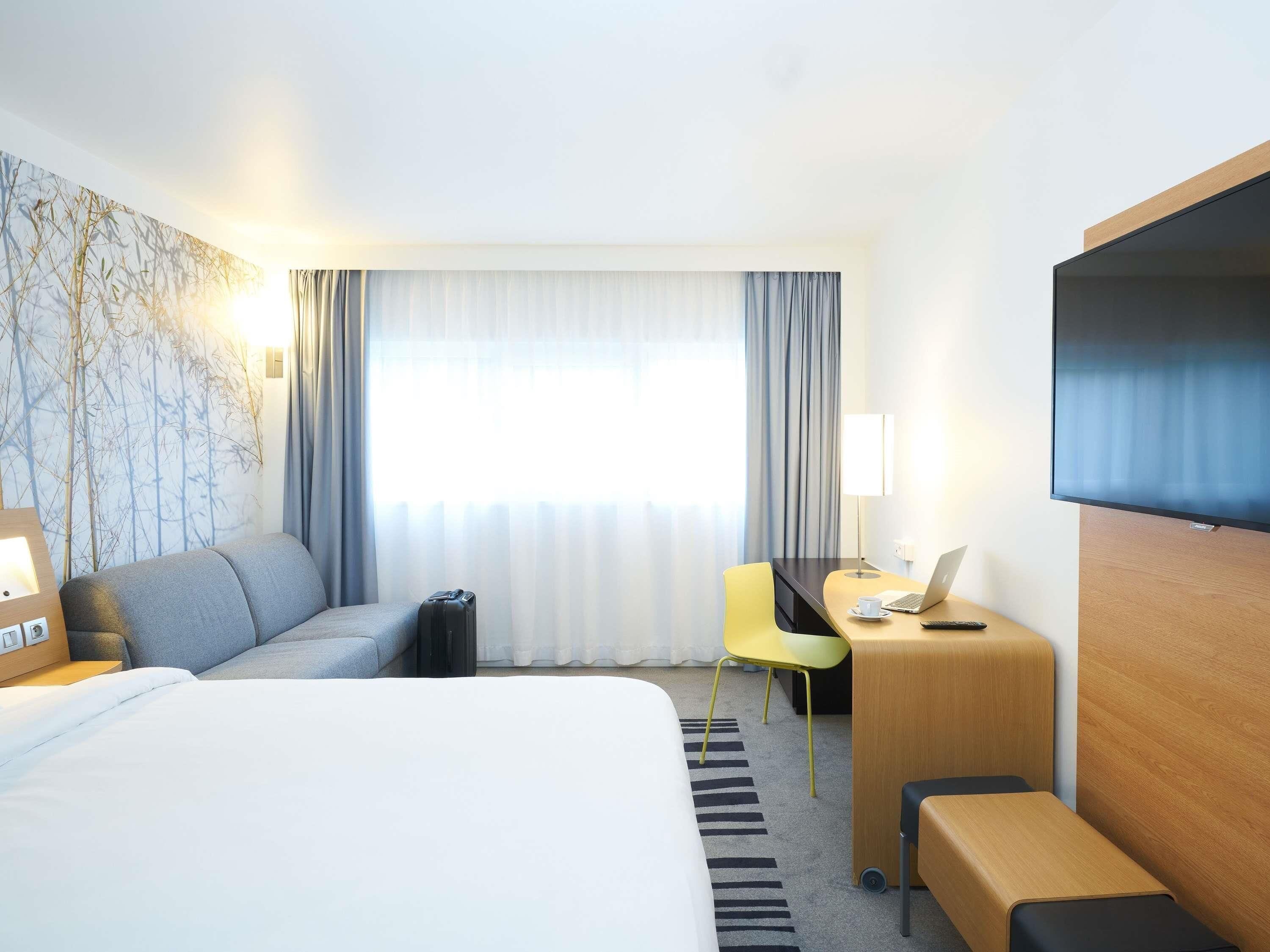 Quarto de hotel com uma cama, uma mesa com um laptop, uma cadeira, uma televisão e um painel decorado com tema de natureza.