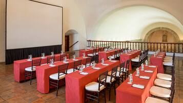 Sala de conferências com mesas longas, cadeiras e uma tela grande.