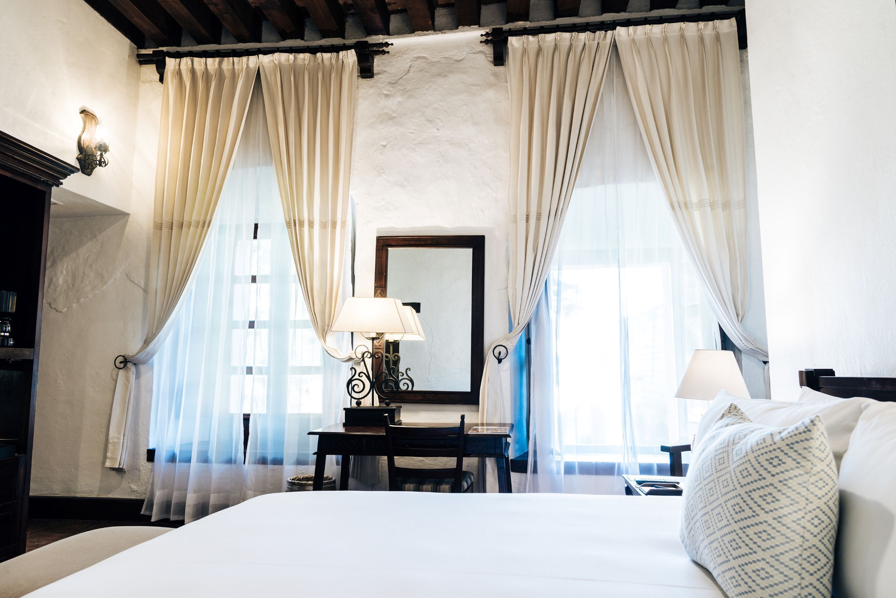 Quarto de hotel com uma cama, uma escrivaninha com luminária, um espelho e cortinas finas.