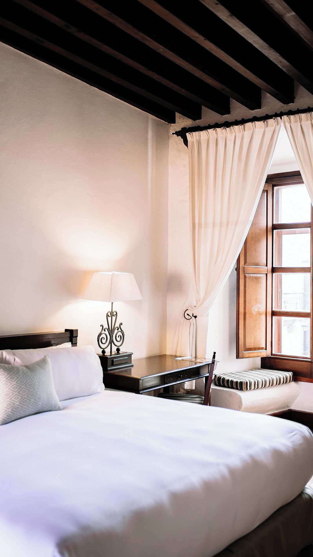 Quarto de hotel com cama, mesa de cabeceira, abajur e janela com cortinas.