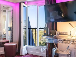 Quarto de hotel moderno com uma janela grande, televisão de tela plana, máquina de café e mesa de vidro com toalha de renda.