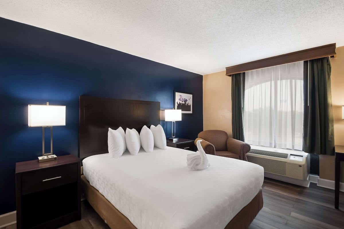 Quarto de hotel com uma cama grande, duas mesas de cabeceira com luminárias, uma cadeira, uma janela com cortinas e um quadro na parede.