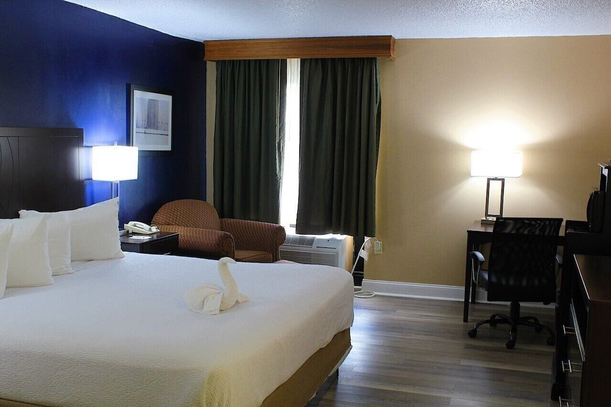 Quarto de hotel com uma cama, duas cadeiras, uma mesa de trabalho, um abajur e uma janela com cortinas.