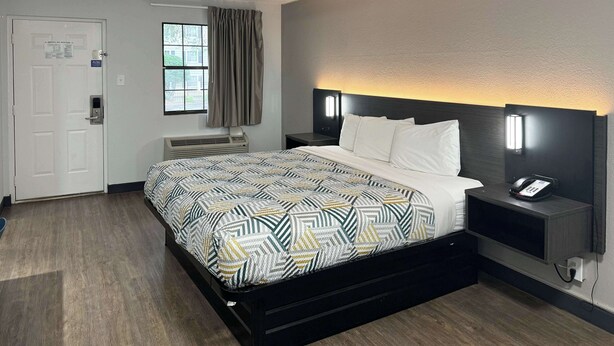 Quarto de hotel com uma cama, uma mesa de cabeceira com telefone, uma luminária na parede e uma janela com cortinas.