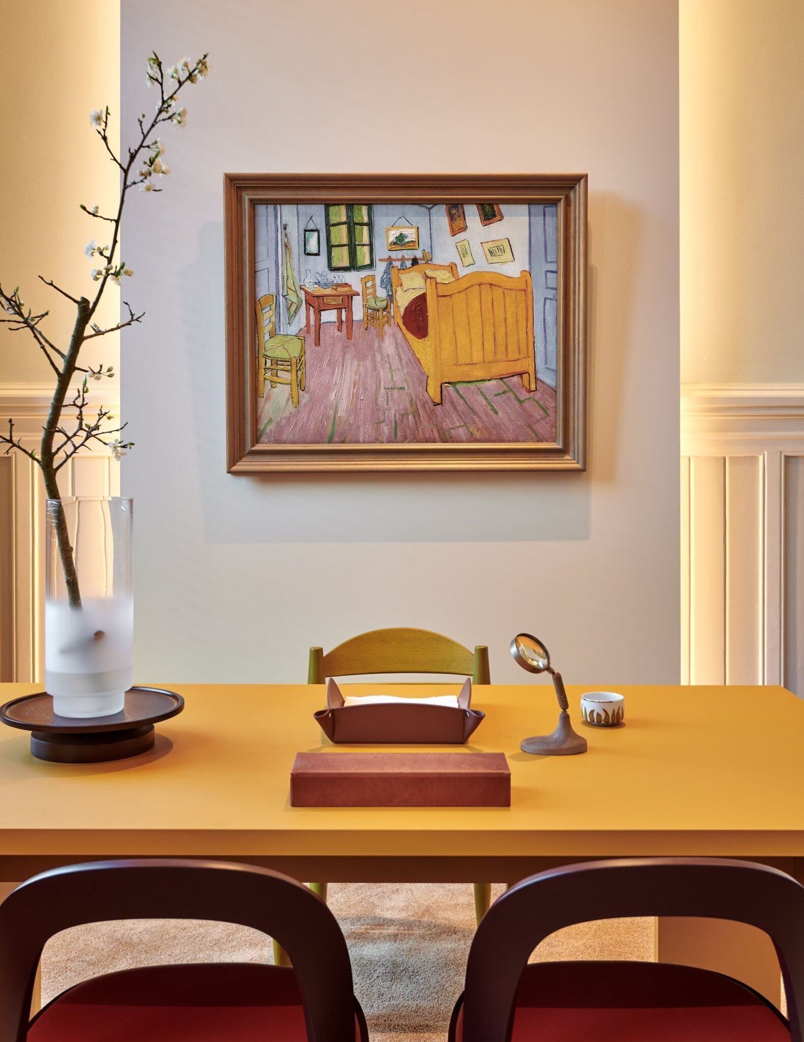 Uma sala de jantar com uma mesa, cadeiras, um vaso com galhos, um quadro emoldurado e um abajur.
