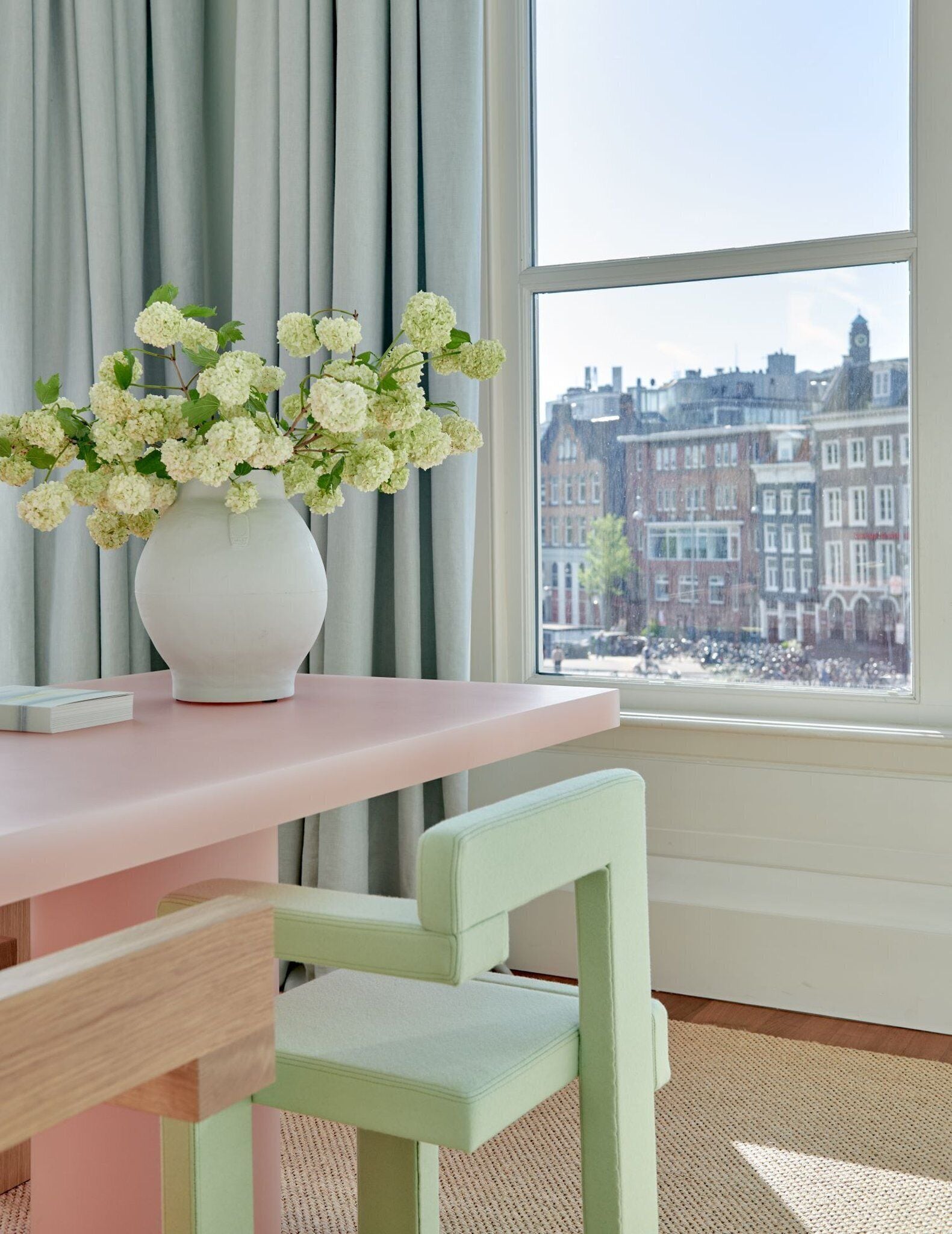 Quarto com uma mesa rosa, um vaso branco com flores, uma cadeira verde e vista para prédios pela janela.