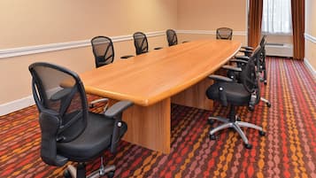 Sala de conferências com uma longa mesa de madeira rodeada por seis cadeiras de escritório pretas com encosto em rede. O espaço possui carpete estampado e paredes bege.