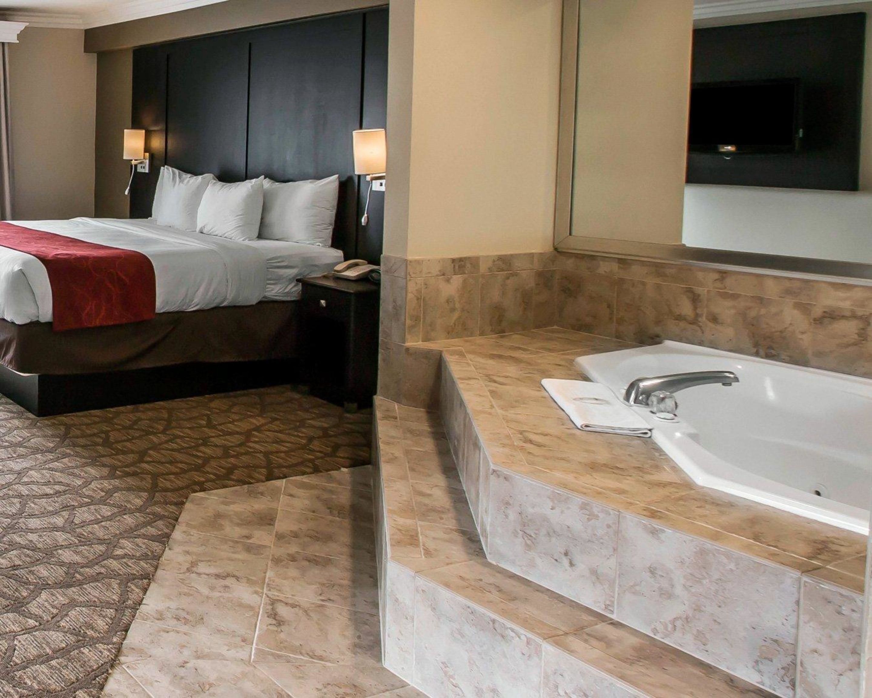 Quarto de hotel com cama, mesa de cabeceira, luminária e banheiro com banheira e pia.