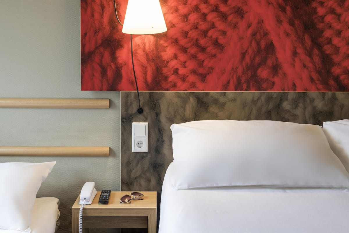 Quarto de hotel com uma cama, uma mesa de cabeceira com telefone e óculos de sol, uma luminária na parede e uma parede vermelha com textura.