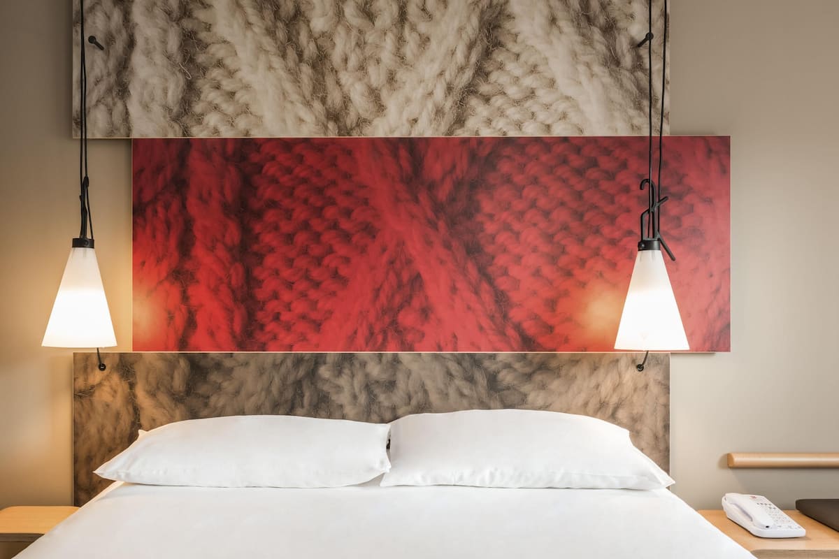 Quarto de hotel com cama, duas luminárias pendentes, quadro de parede texturizado em vermelho e bege, travesseiro branco e mesa de cabeceira com telefone.