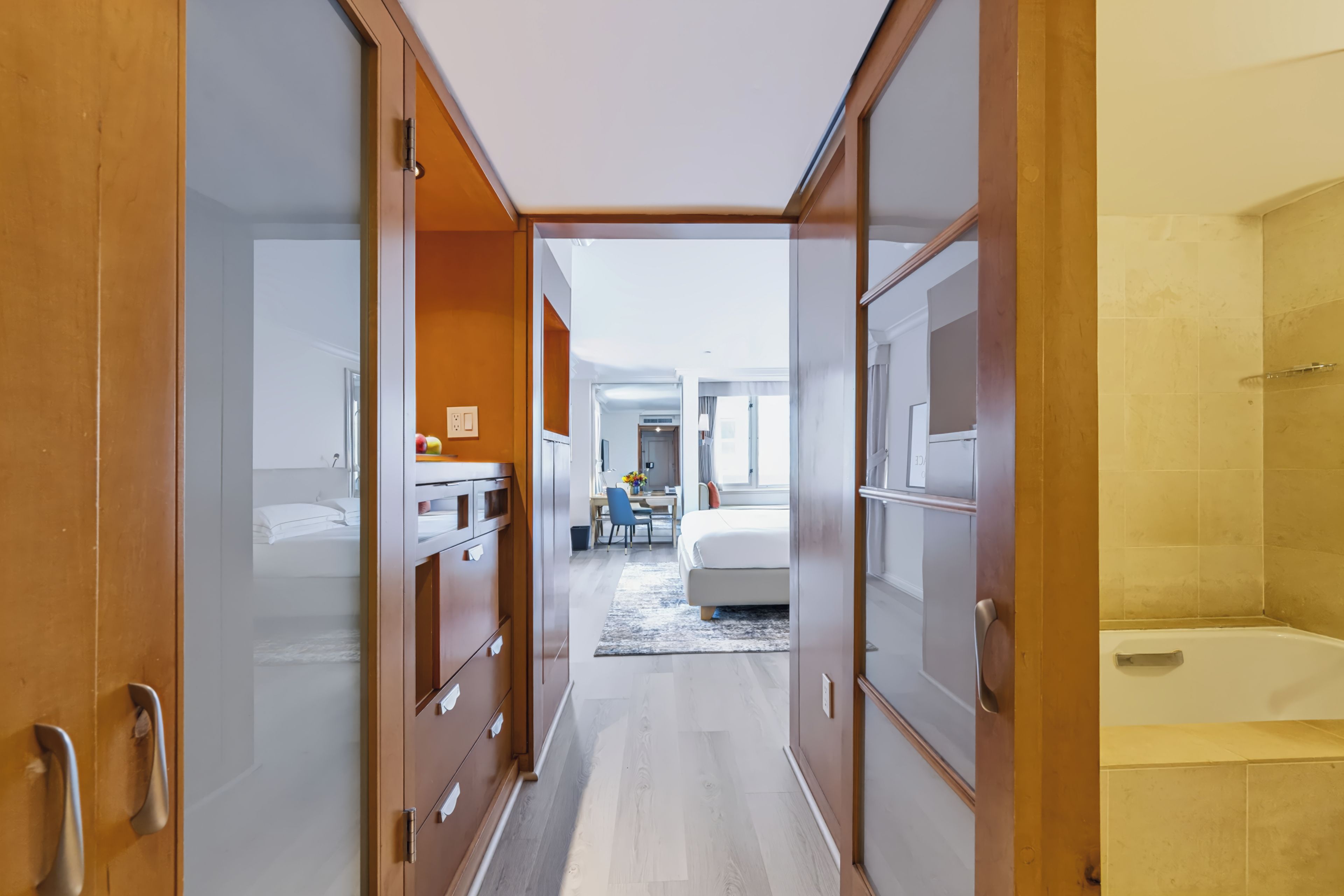 Corredor que leva a um quarto com uma cama, uma escrivaninha e uma cadeira. Banheiro com banheira visível à direita.