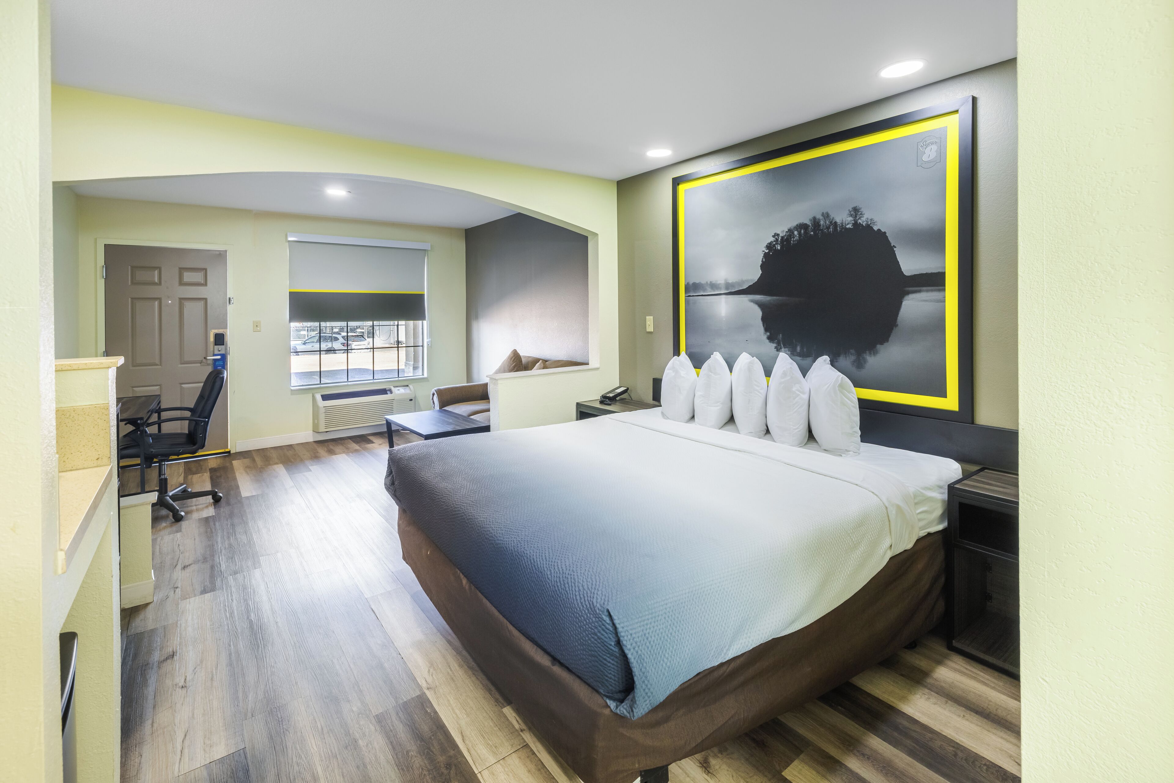 Quarto de hotel com uma cama grande, uma escrivaninha, uma cadeira, uma janela com persianas e um quadro de um lago.