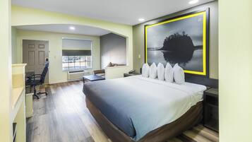 Quarto de hotel com uma cama grande, uma escrivaninha, uma cadeira, uma janela com persianas e um quadro de um lago.