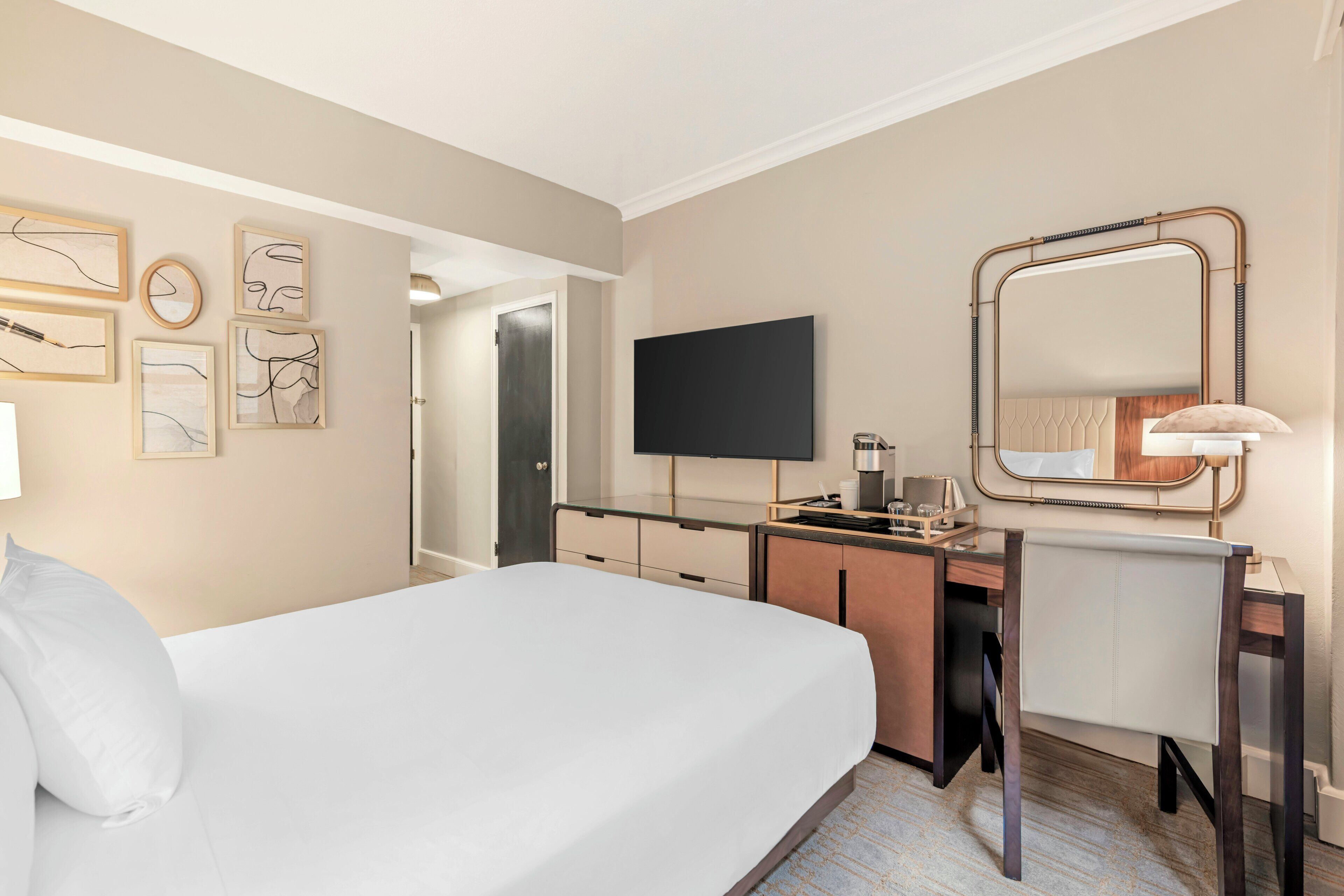 Quarto de hotel com cama, escrivaninha, televisão de tela plana, espelho e obras de arte nas paredes.