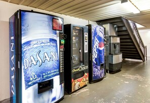 Uma fileira de máquinas de venda automática com opções de água e bebidas, incluindo Dasani e Pepsi, em um espaço com piso de concreto e uma escada ao fundo.