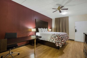 Quarto de hotel com cama, mesas de cabeceira, escrivaninha, ventilador de teto e porta.