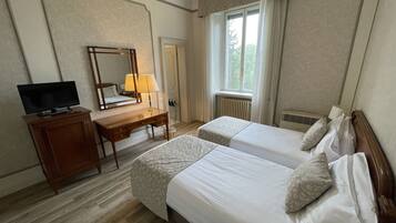 Quarto de hotel com duas camas, uma escrivaninha, televisão, espelho e janela com vista para árvores.