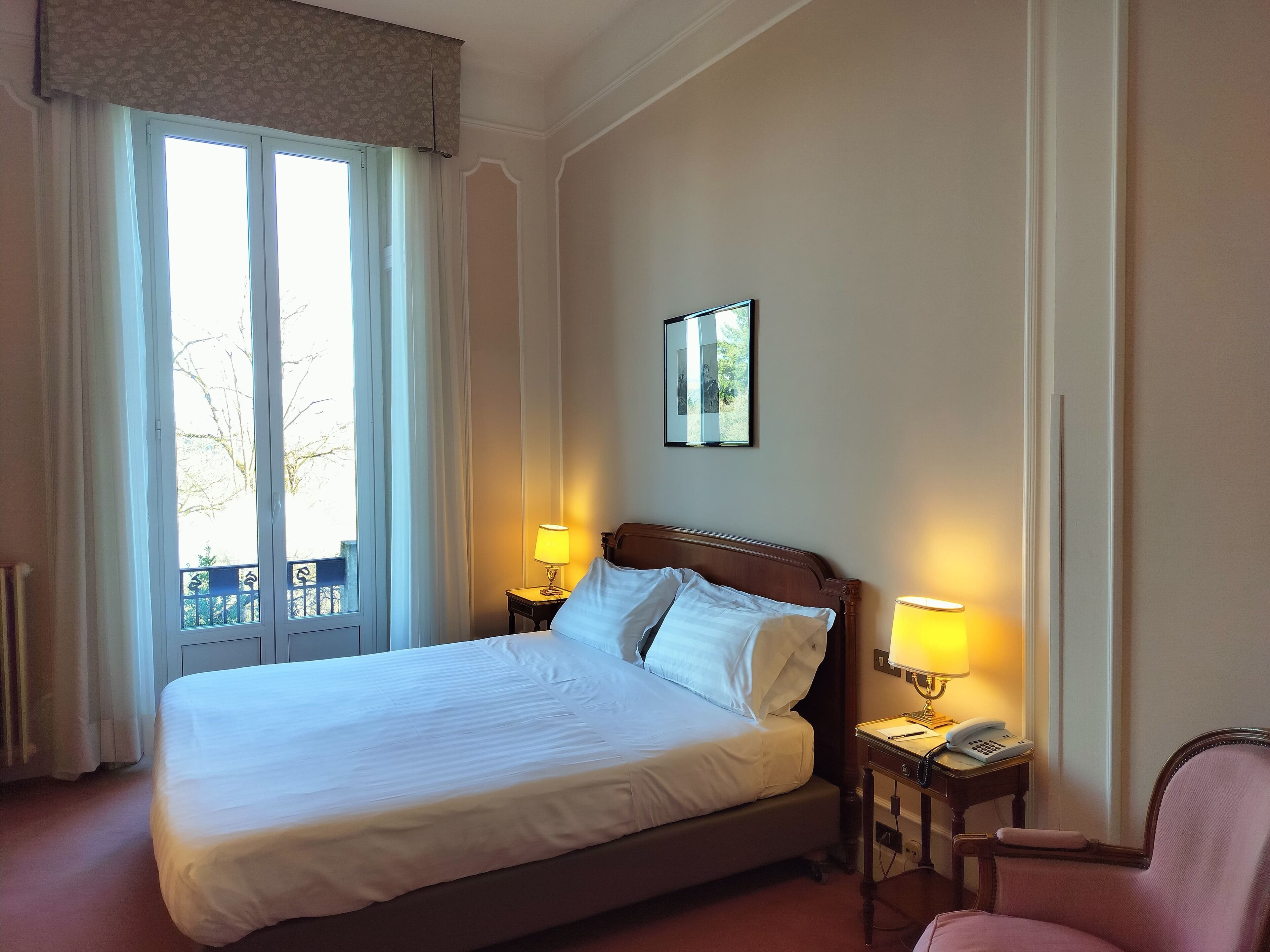 Quarto de hotel com cama grande, duas luminárias de cabeceira, mesa de apoio com telefone e uma cadeira.