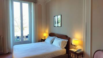Quarto de hotel com cama grande, duas luminárias de cabeceira, mesa de apoio com telefone e uma cadeira.