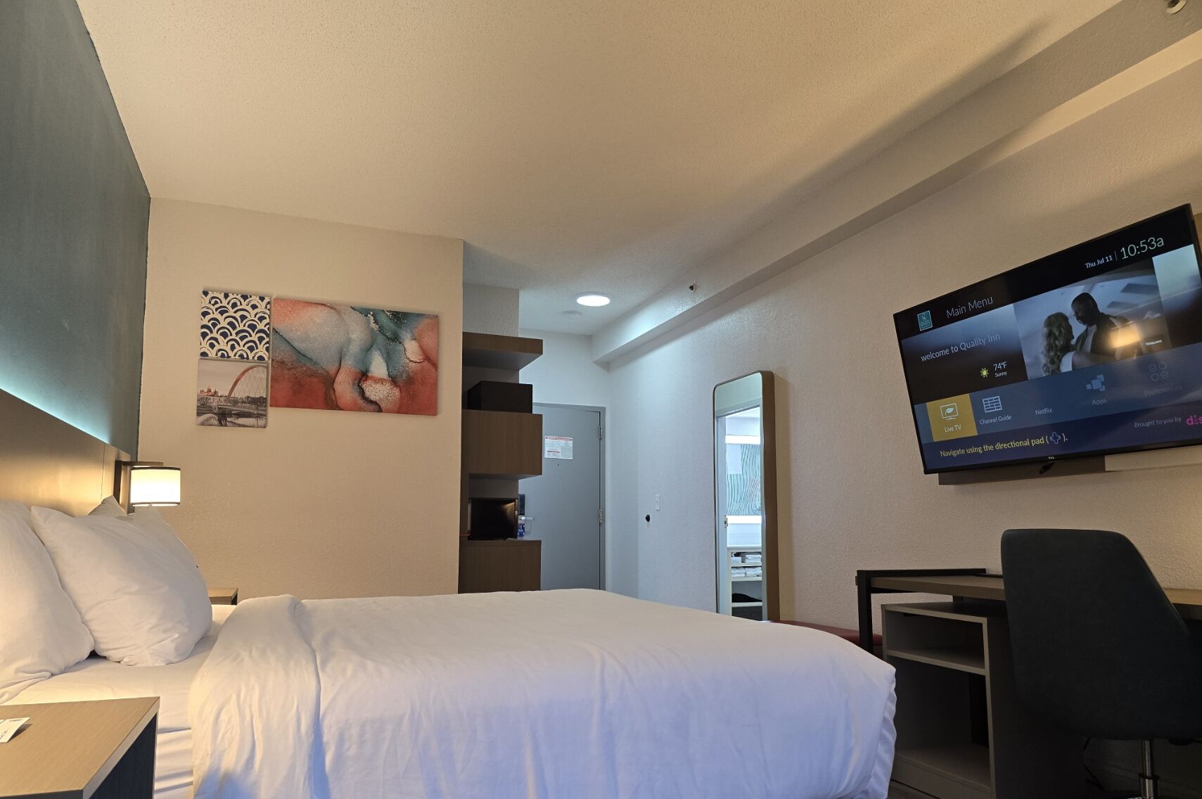 Quarto de hotel com cama, escrivaninha, cadeira, TV presa na parede e obras de arte na parede.