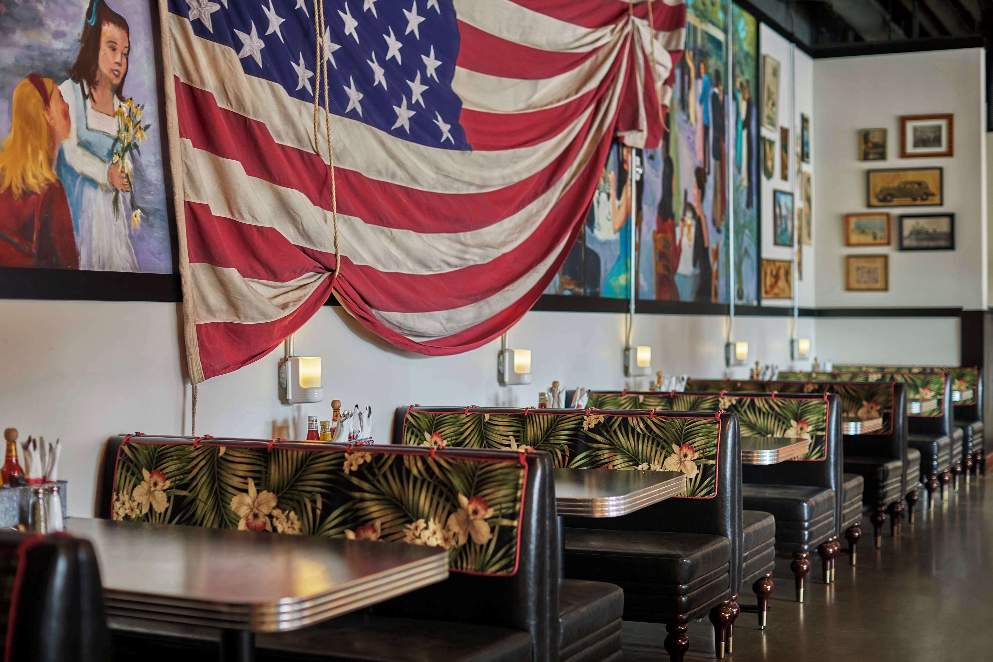 Interior de restaurante com decorações de bandeira americana, cabines com tema tropical e obras de arte emolduradas nas paredes.