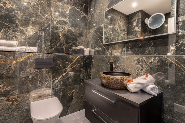 Um banheiro moderno com painel de mármore, uma pia redonda com design exclusivo, vaso sanitário e bancada com gavetas.