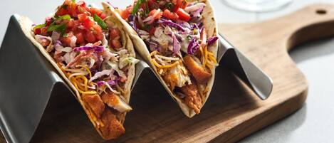Dois tacos sobre uma tábua de madeira, com uma taça de vinho ao fundo.