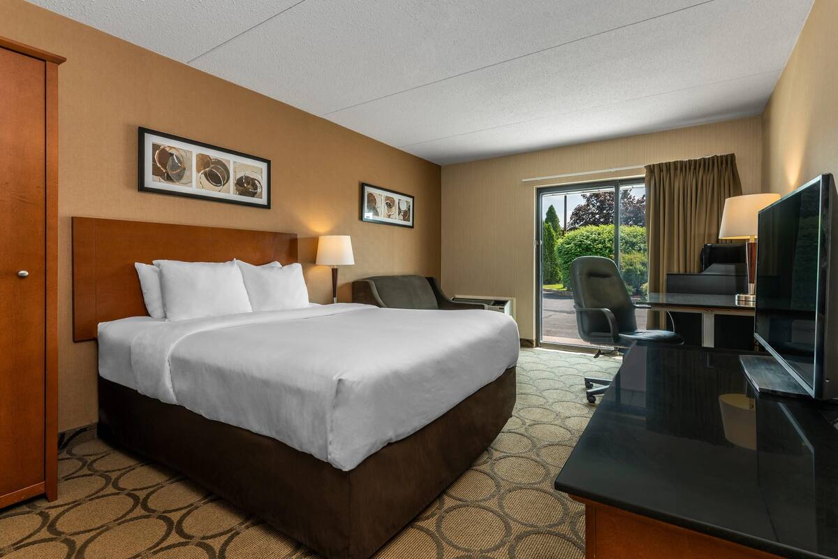 Quarto de hotel com uma cama grande, uma escrivaninha com televisão, uma cadeira e uma janela com cortinas.