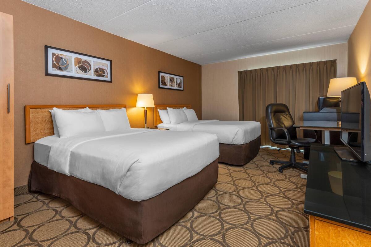 Quarto de hotel com duas camas, uma escrivaninha, uma cadeira, televisão e obras de arte nas paredes.