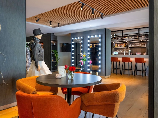 Interior moderno com mesa redonda, cadeiras laranja, área de bar com banquetas e um manequim vestido de preto.