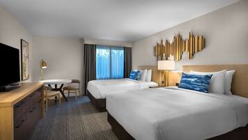 Quarto de hotel com duas camas, uma escrivaninha, uma cadeira, televisão e uma janela grande com cortinas.