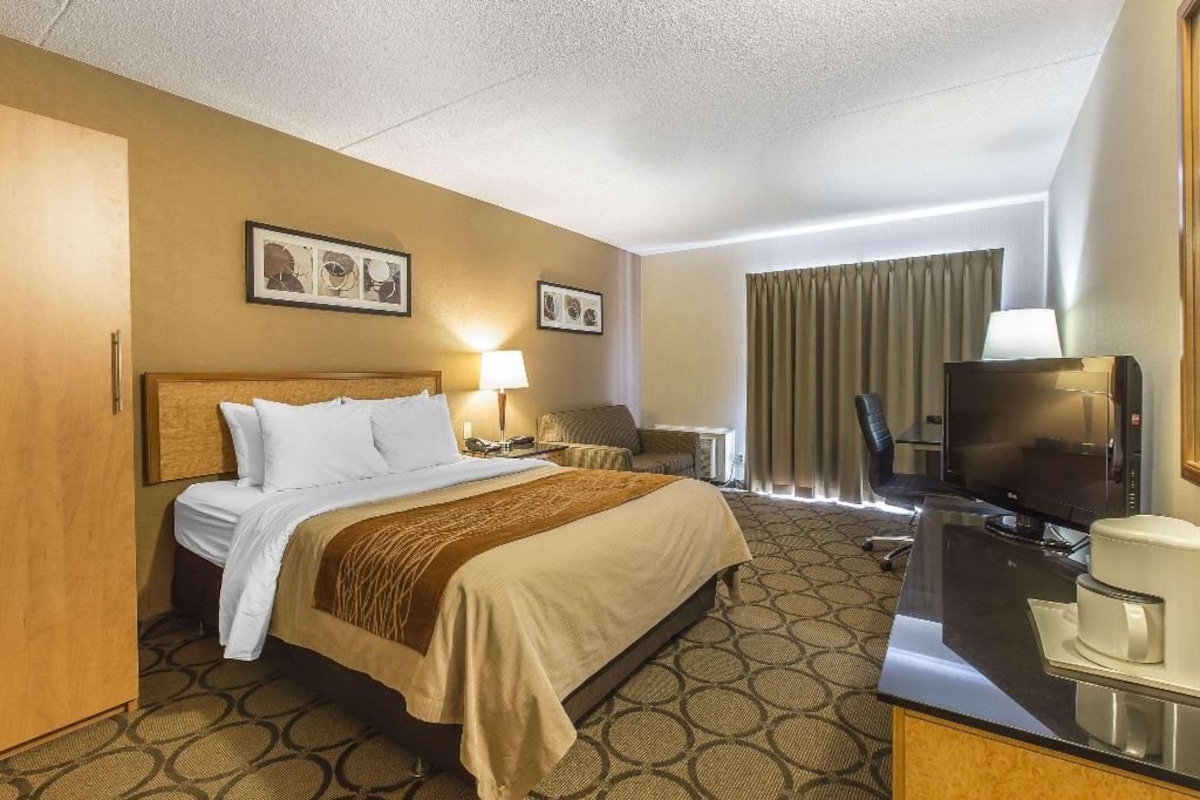 Quarto de hotel com cama, escrivaninha com computador, cadeira e televisão.