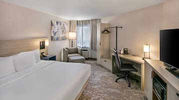 Quarto de hotel com cama, escrivaninha, cadeira, televisão, luminária e uma obra de arte na parede.