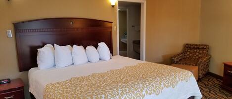 Quarto de hotel com uma cama grande, cabeceira de madeira, duas luminárias de parede, tapete estampado, uma poltrona pequena e uma mesa de cabeceira com telefone.