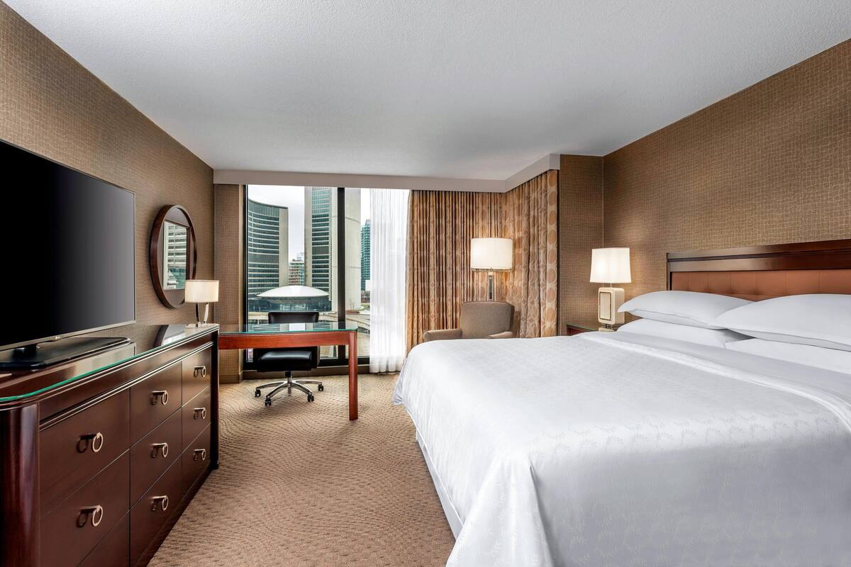 Quarto de hotel com uma cama grande, televisão, escrivaninha com cadeira e vista para a cidade pela janela.
