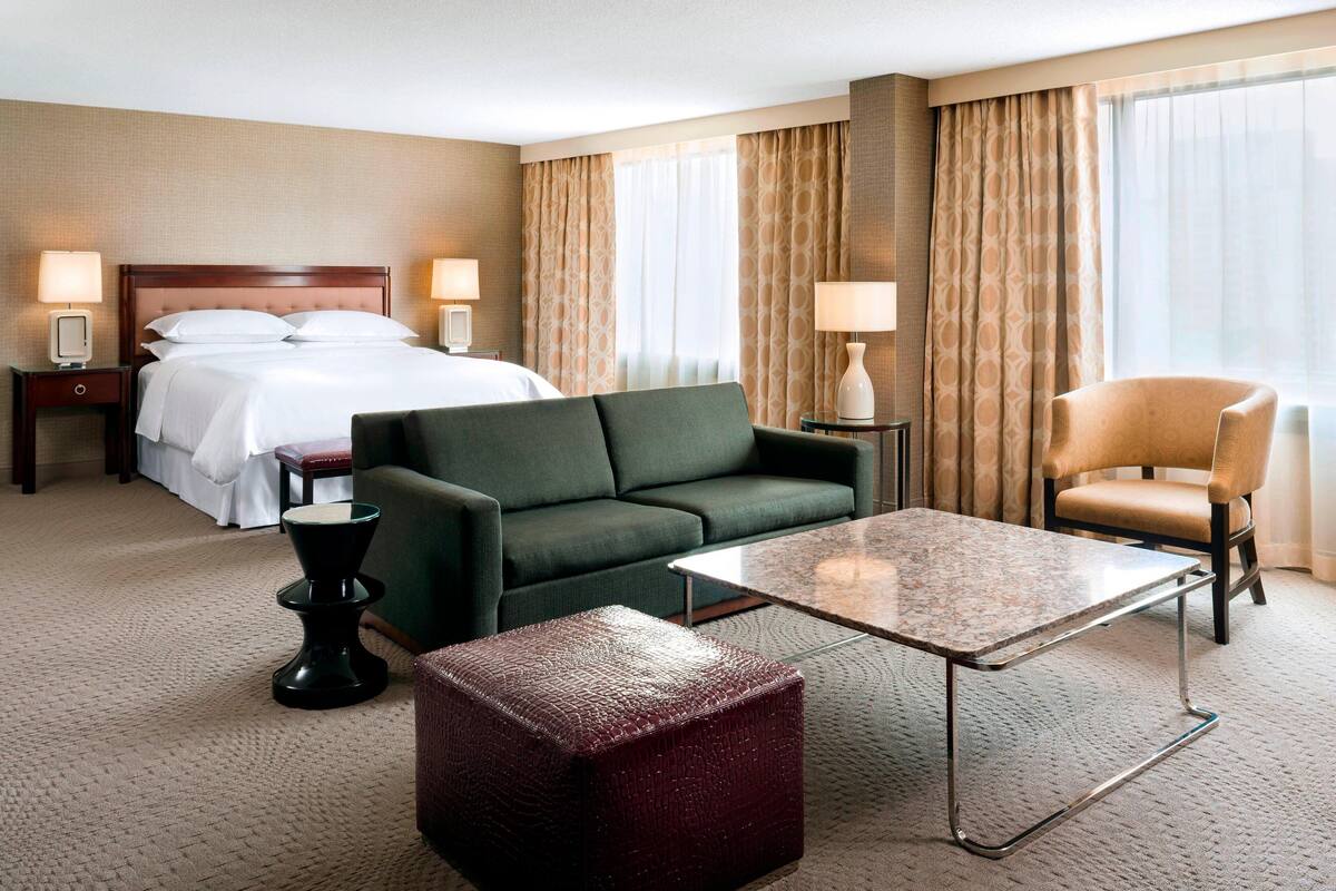 Quarto de hotel com cama, sofá verde, poltrona, mesa de centro e mesinha de cabeceira.