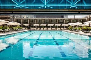 Uma piscina com espreguiçadeiras e guarda-sóis, rodeada por um edifício moderno com grandes janelas.