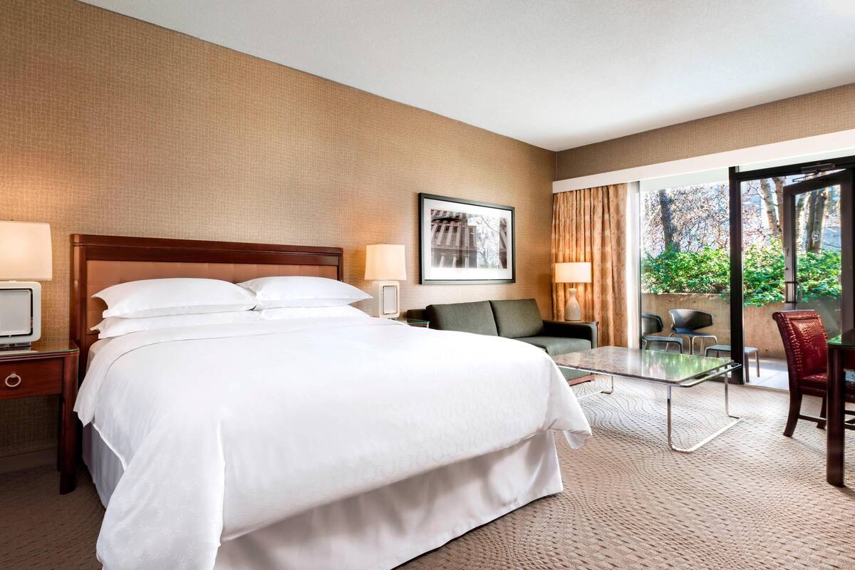 Quarto de hotel com uma cama grande, uma escrivaninha, uma cadeira, uma televisão e vista para árvores pela janela.