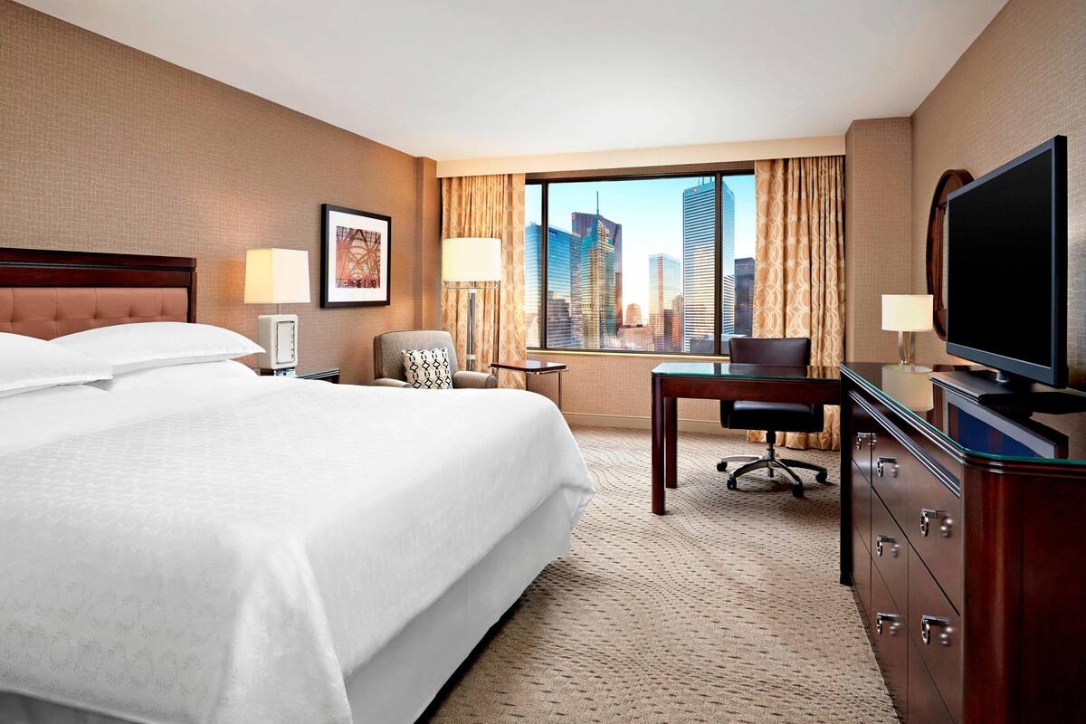 Quarto de hotel com uma cama grande, uma escrivaninha, uma cadeira, televisão e janela com vista para a cidade.