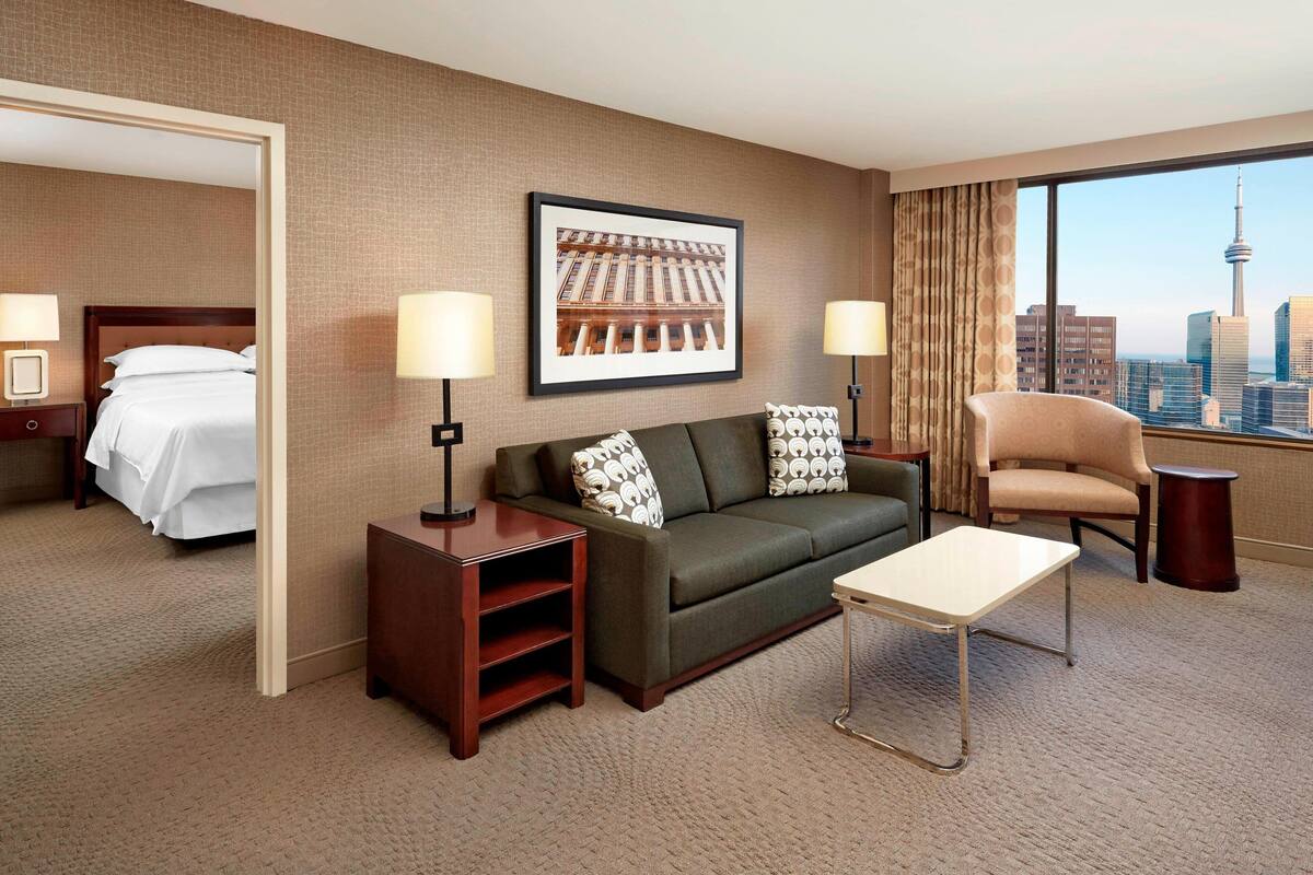 Quarto de hotel com cama, sofá, mesa de centro e vista para o horizonte da cidade.