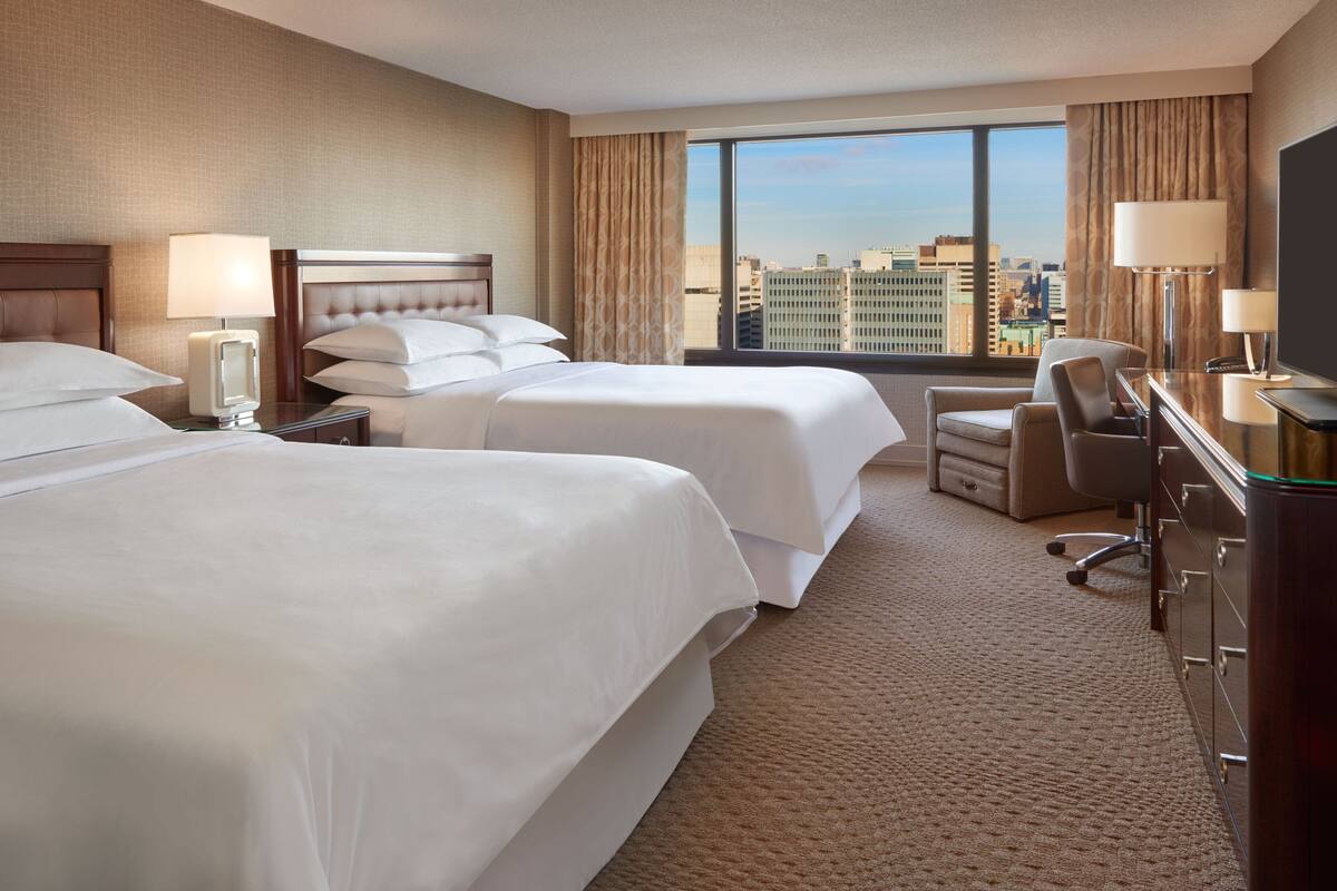 Quarto de hotel com duas camas, uma escrivaninha, uma cadeira e uma janela grande com vista para a cidade.
