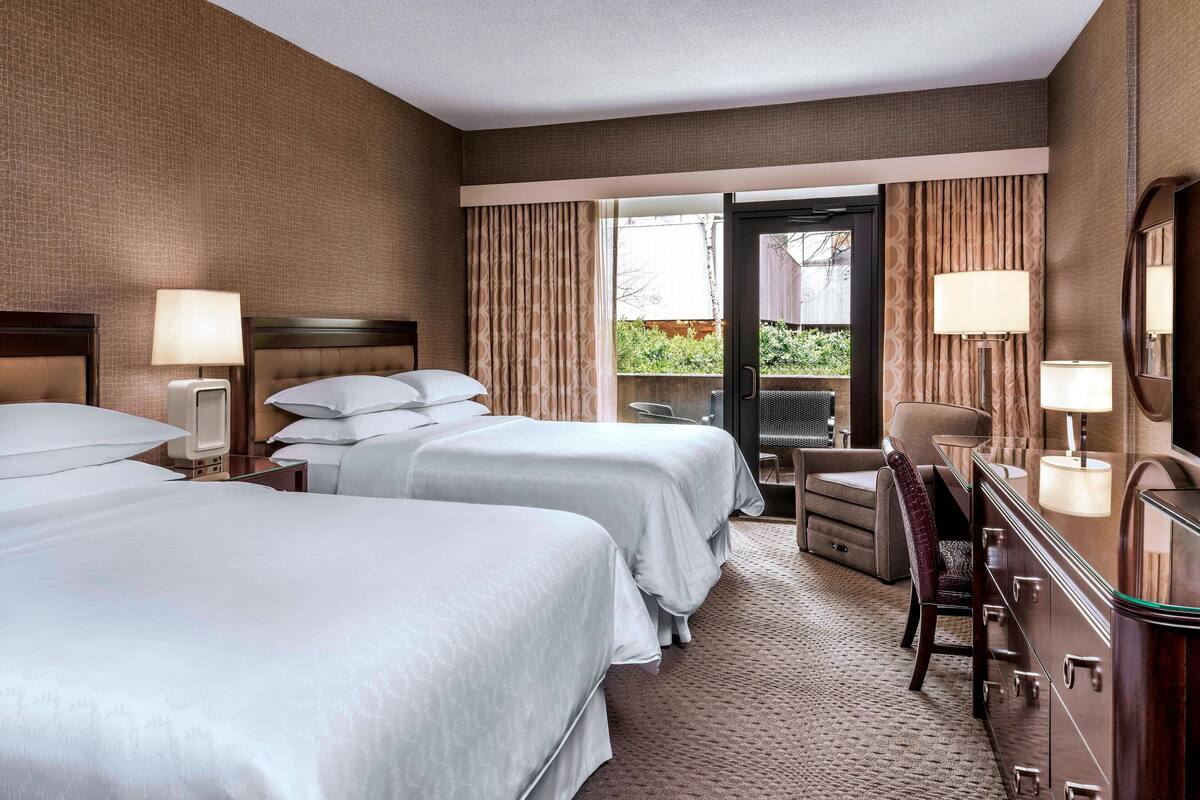 Quarto de hotel com duas camas, uma escrivaninha, uma cadeira, um armário e uma janela com cortinas.