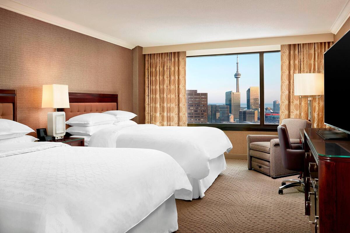 Quarto de hotel com duas camas, uma escrivaninha, uma cadeira e uma janela grande com vista para a paisagem urbana.