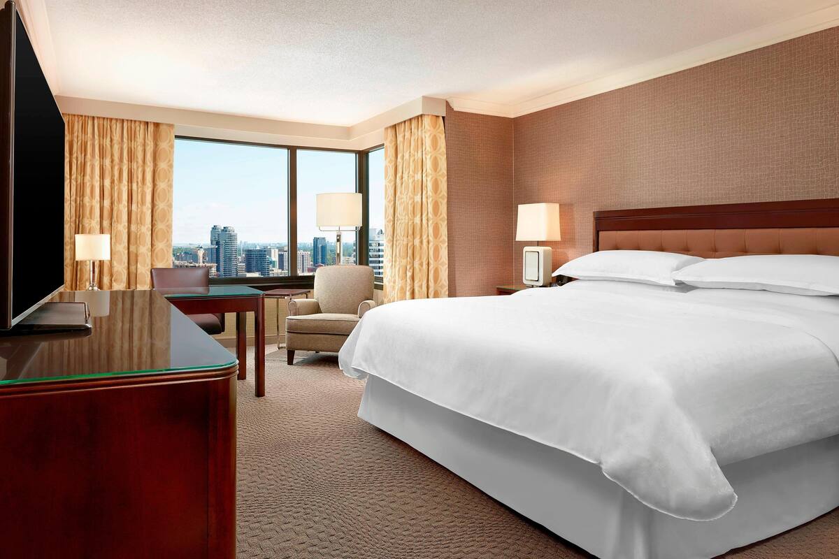 Quarto de hotel com uma cama grande, uma secretária com televisão, uma cadeira e vista da cidade por grandes janelas.