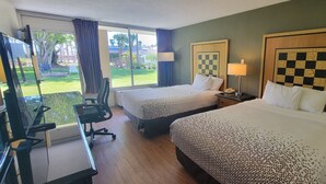Quarto de hotel com duas camas, uma escrivaninha com computador e uma janela grande com vista para um jardim e árvores.
