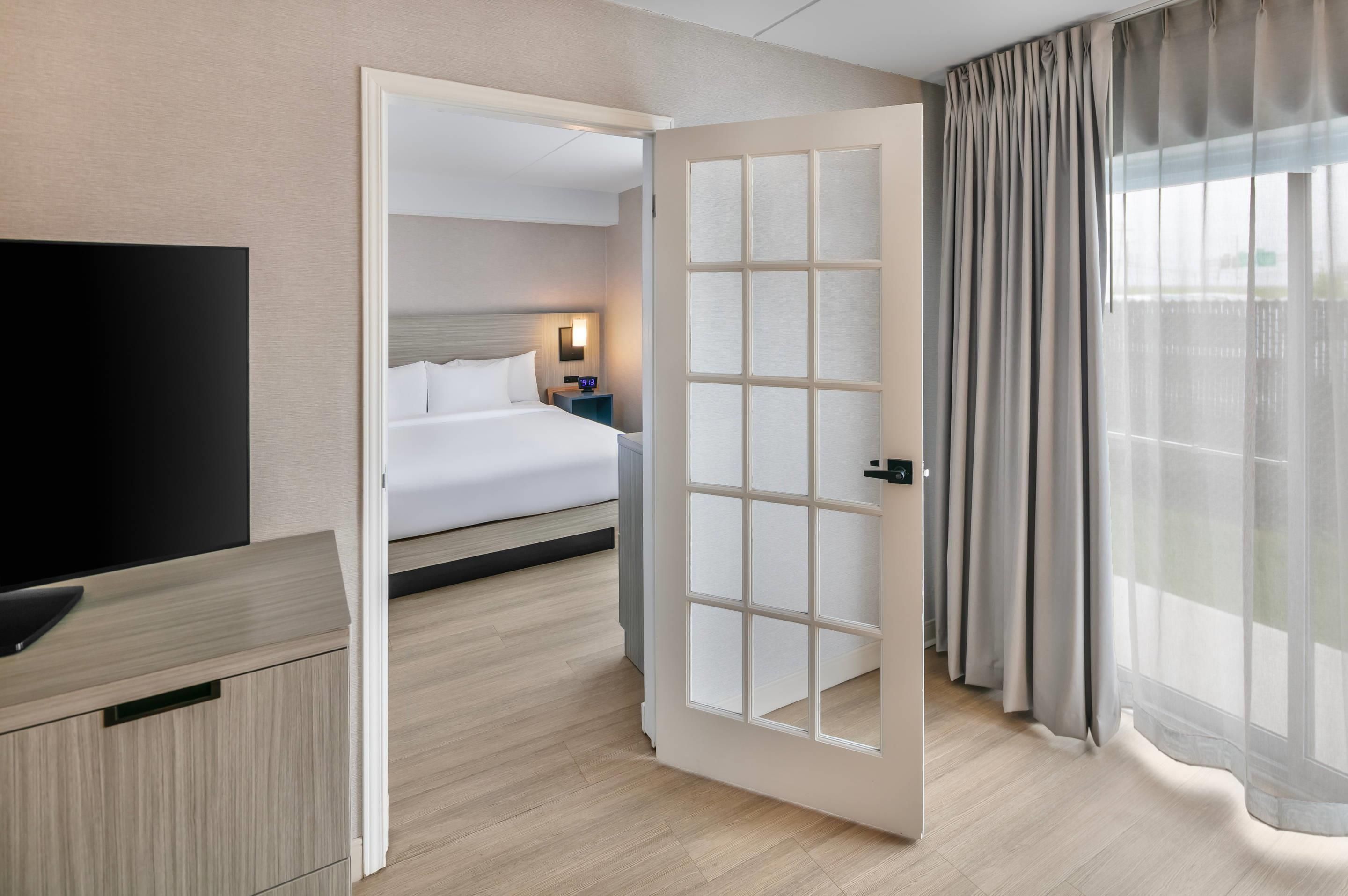 Quarto de hotel com cama, televisão, mesa de cabeceira e porta que dá para outro espaço.
