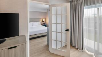 Quarto de hotel com cama, televisão, mesa de cabeceira e porta que dá para outro espaço.
