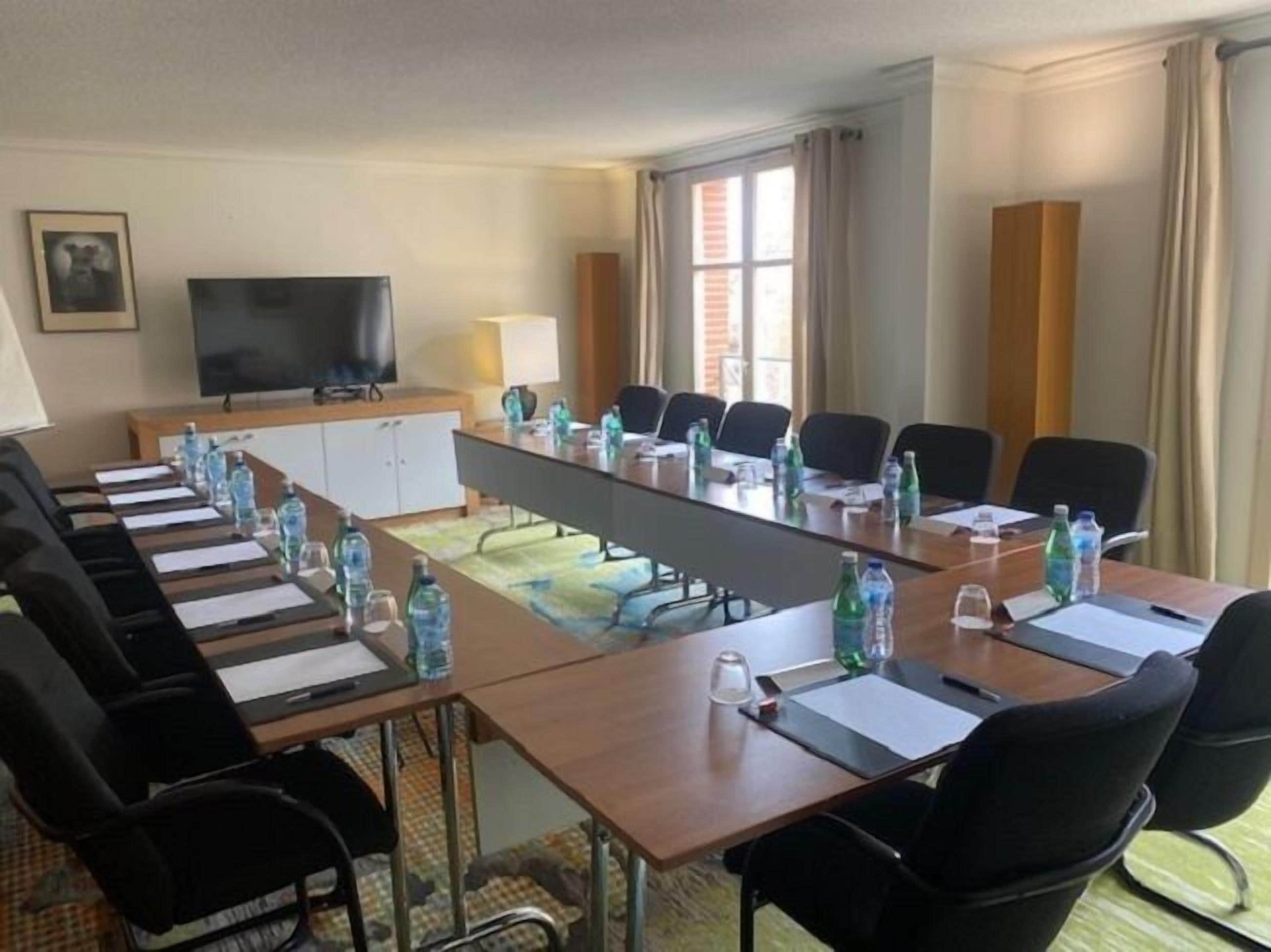 Uma sala de conferência com uma mesa comprida, cadeiras, garrafas de água e uma televisão sobre um suporte.
